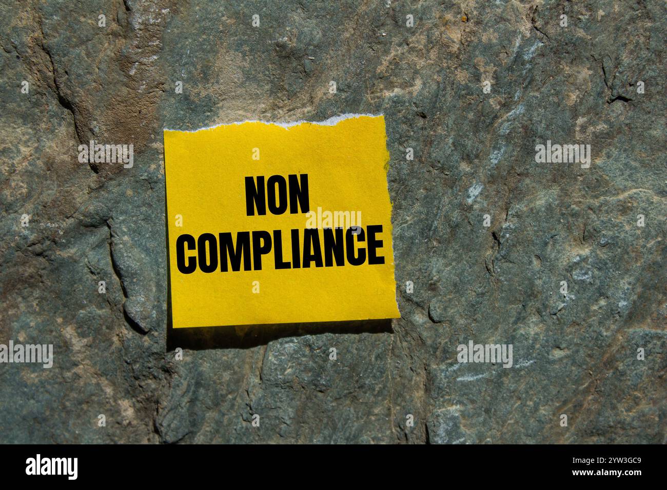 Non compliance message written on ripped torn yellow paper piece with stone background ...
