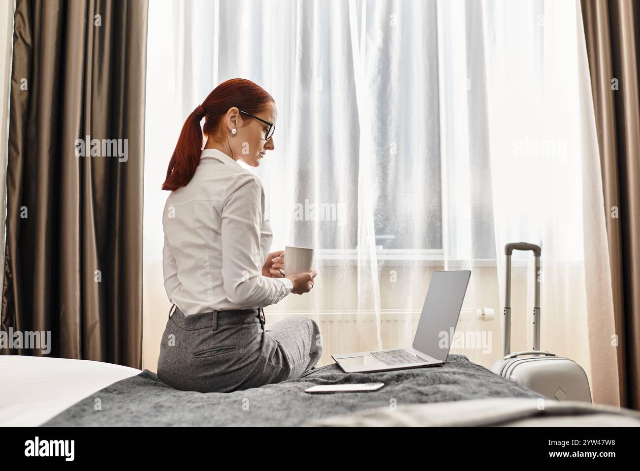 A young woman sits on a bed with a cup of coffee, focused on her laptop ...