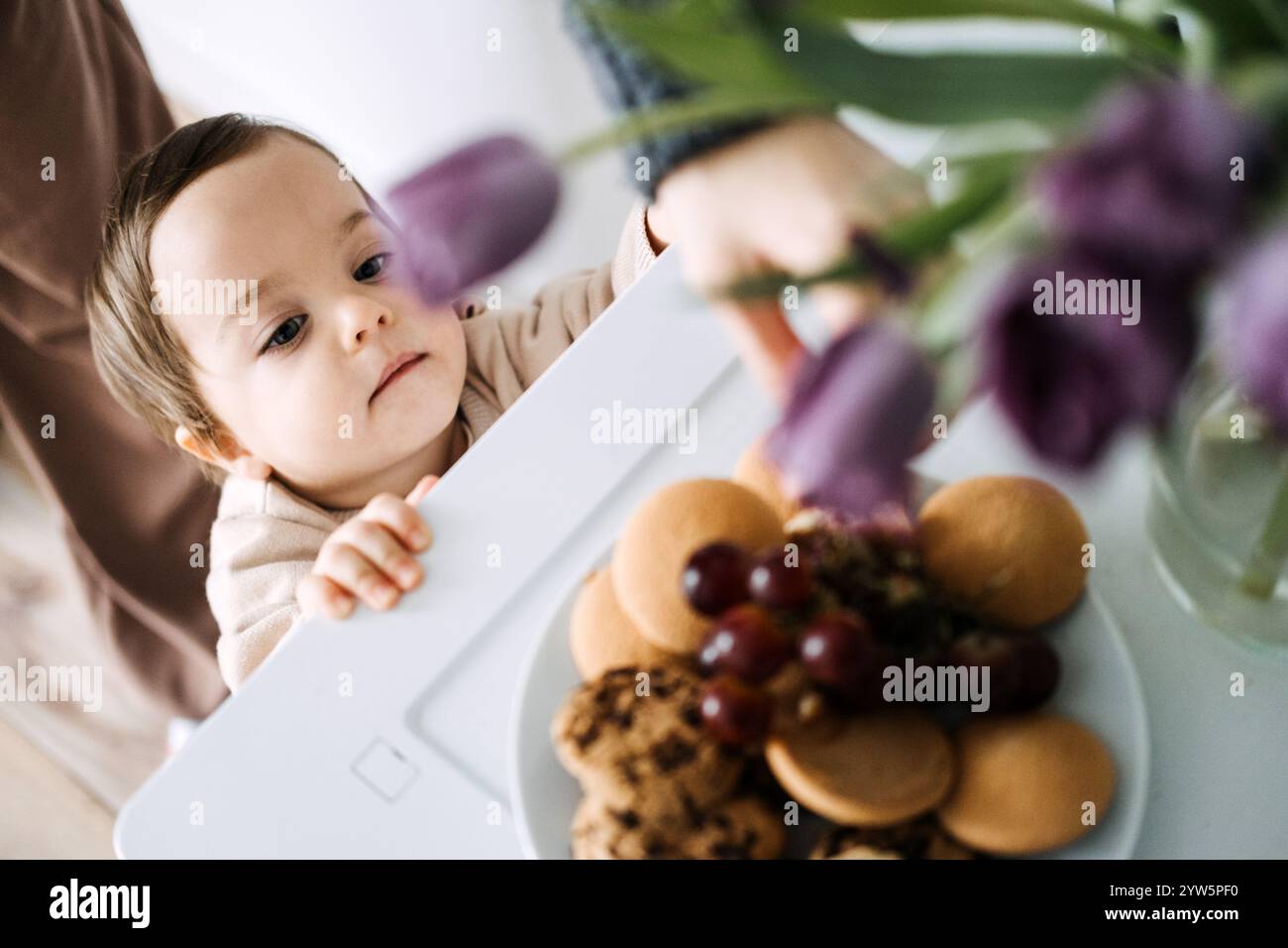 Curious toddler reaching for snacks on a table near flowers. Childhood ...