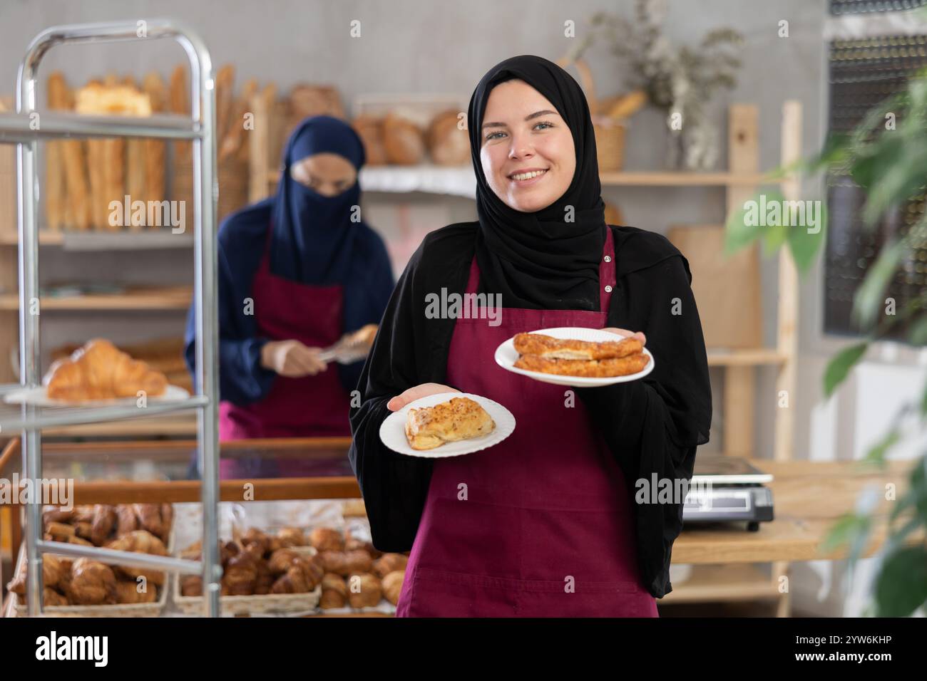 Positive girl seller of oriental bakery in traditional Muslim clothes with fresh hot pastries in ...
