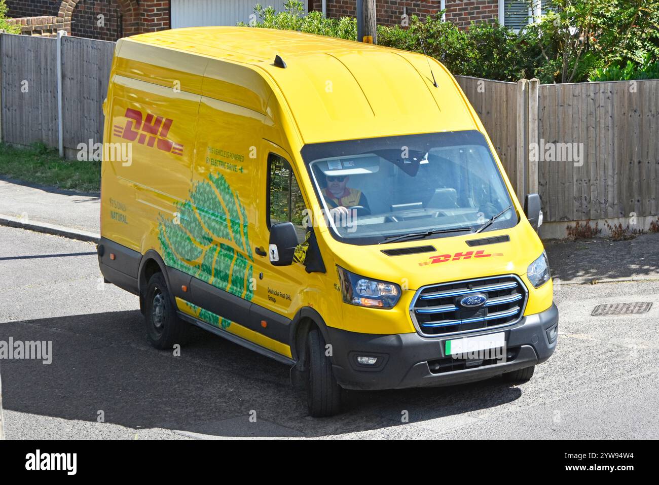 DHL a multinational logistics & parcel delivery brand yellow Ford Transit van driving on village ...