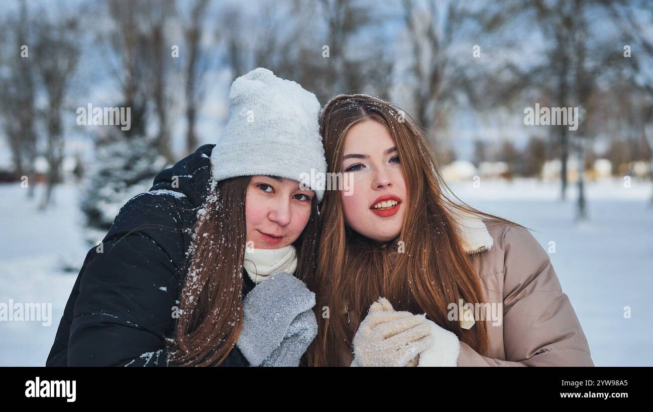 Two young girls in winter attire are hugging in a snowy park, savoring ...