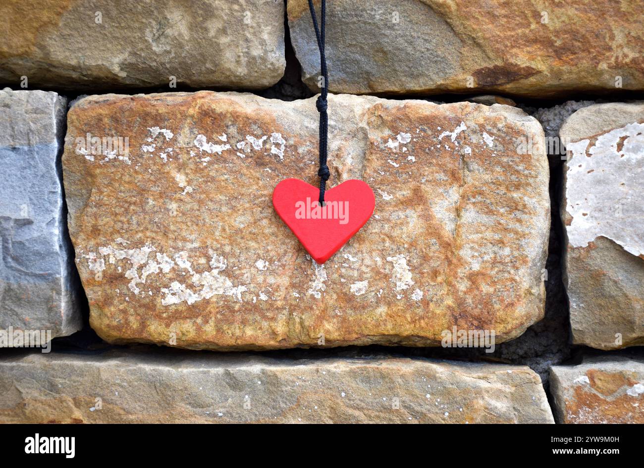 A red heart-shaped pendant hanging from a black string against a stone ...