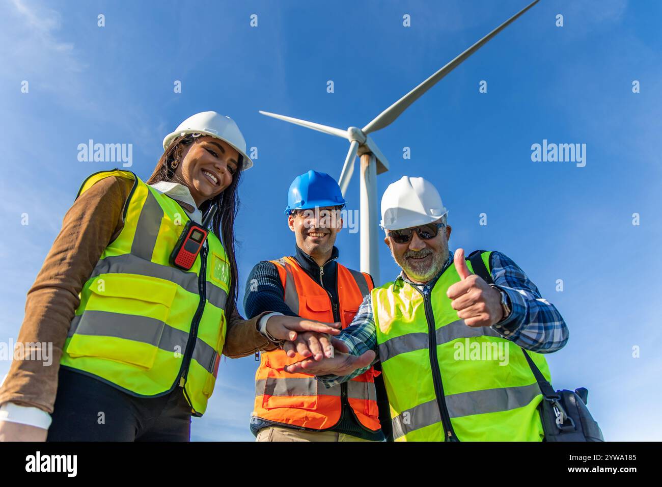 Three multiethnic engineers in high-visibility vests and hard hats joining hands beneath a wind ...