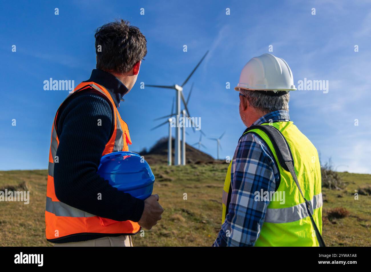 Two engineers are inspecting a wind turbine farm on a hilltop, wearing ...
