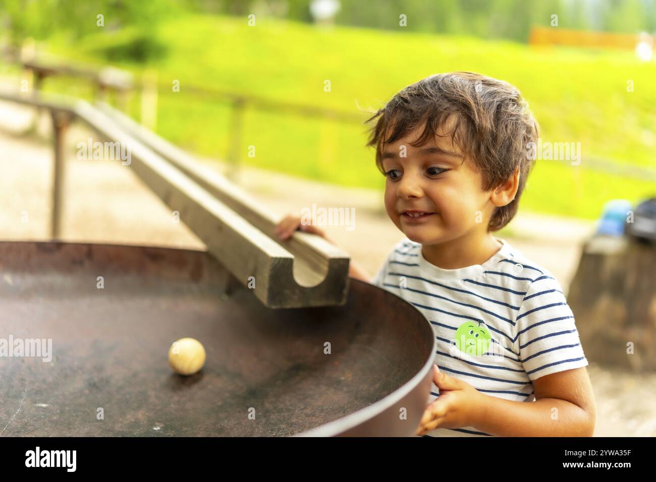 Curious child having fun with a wooden ball rolling down a track in a playground, learning about ...