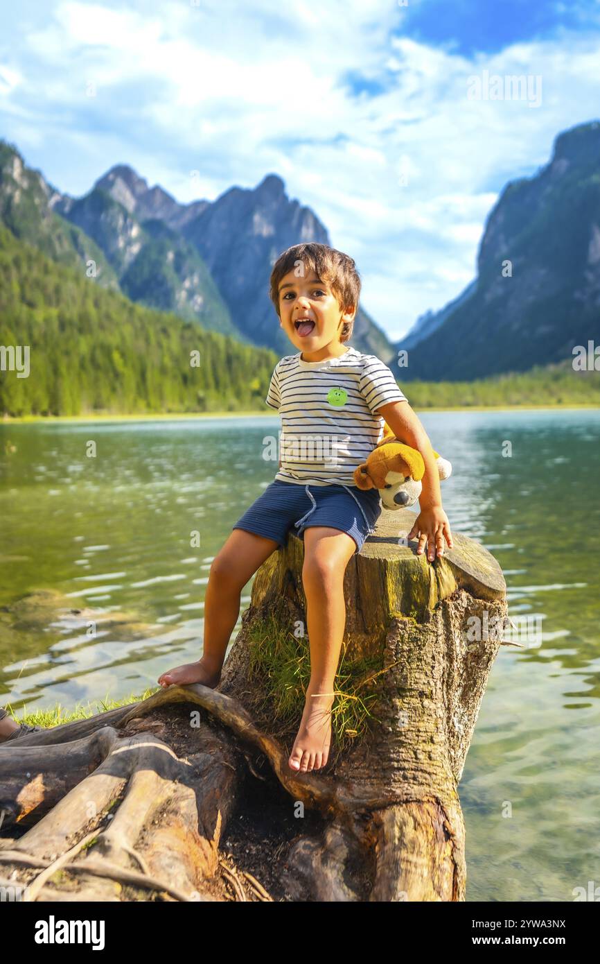 Carefree child sticking out tongue, sitting on tree stump by lake ...