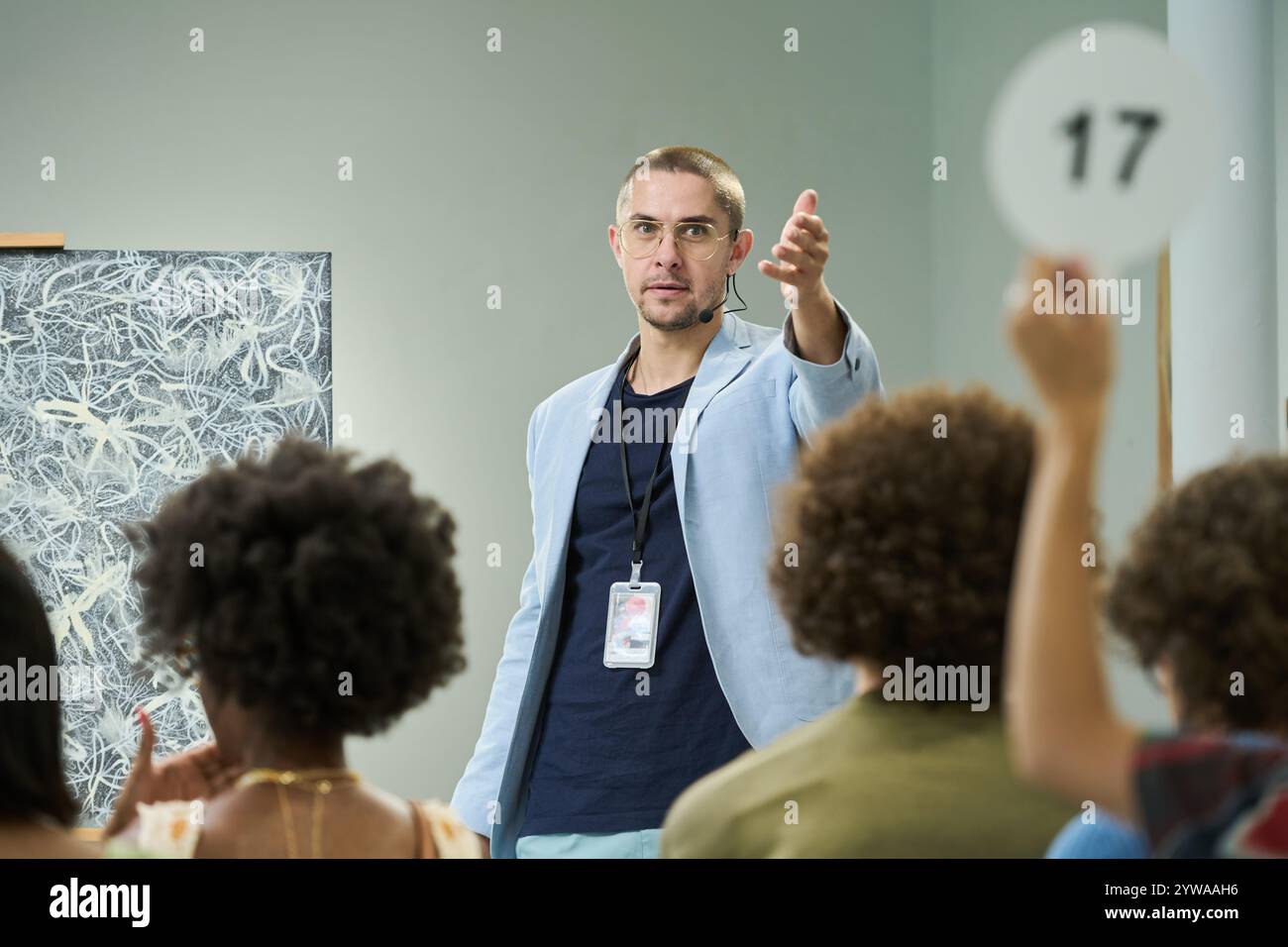 Young elegant man pointing at one of auctioneers raising hand holding ...