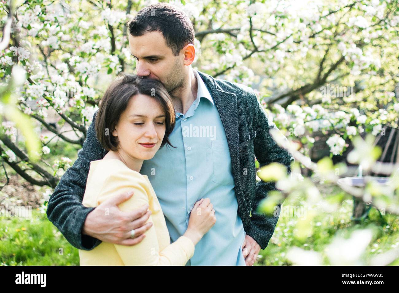 a husband and a pregnant wife hug in a spring garden surrounded by ...