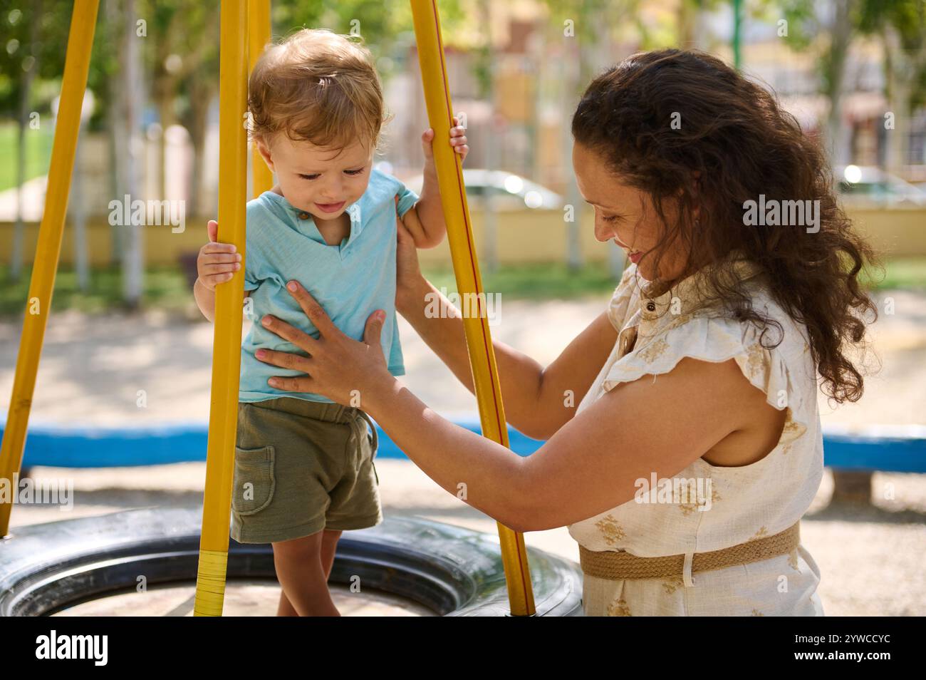 A caring mother helps her child play safely on a swing at a sunny ...