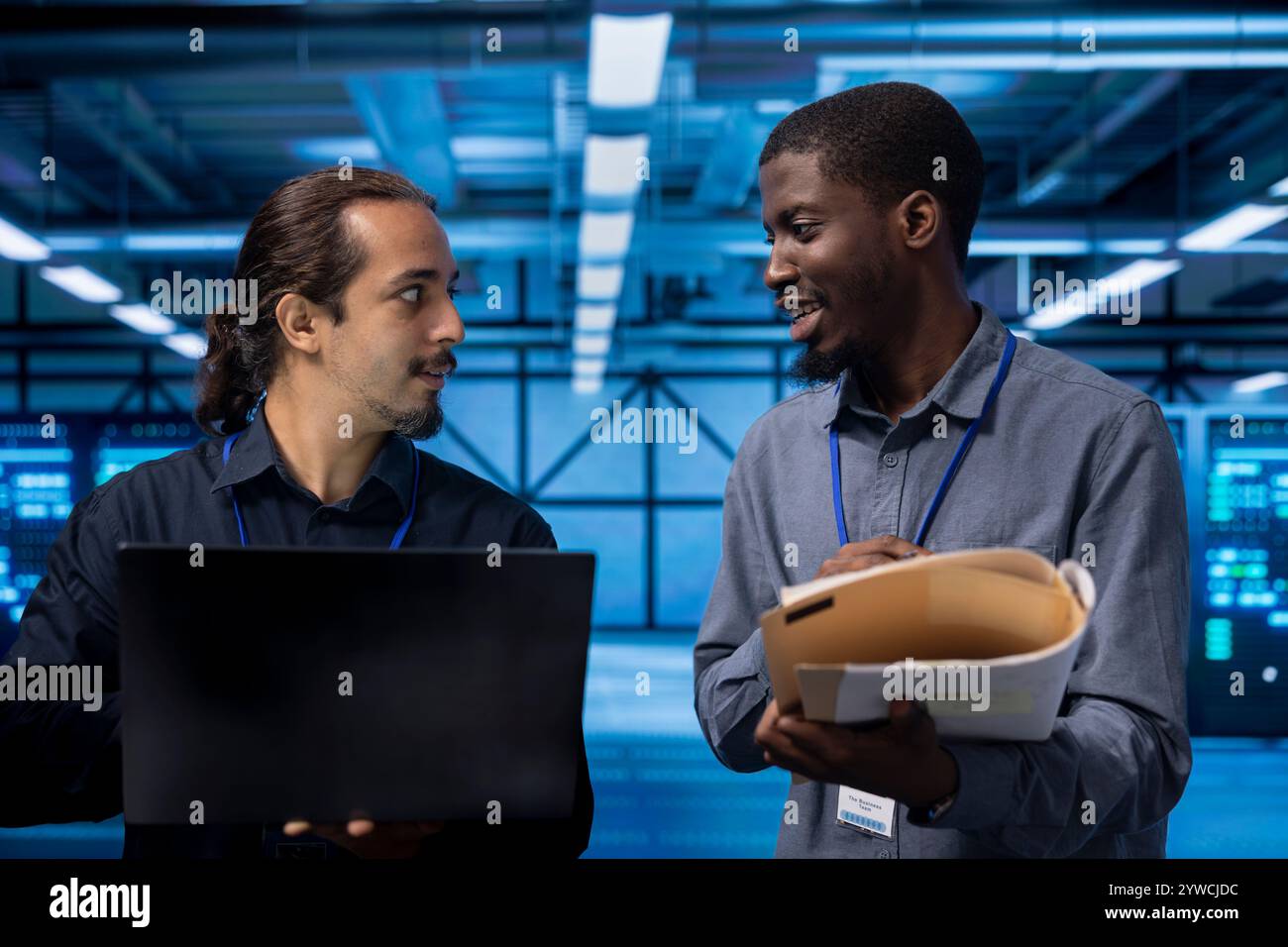 Smiling manager inspecting server room equipment, reading paperwork from folder, talking with ...