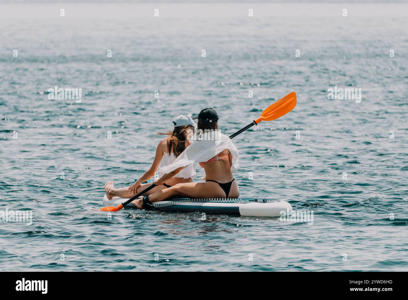 Two women floating on a surfboard in the ocean. One of them is wearing a white shirt and the ...