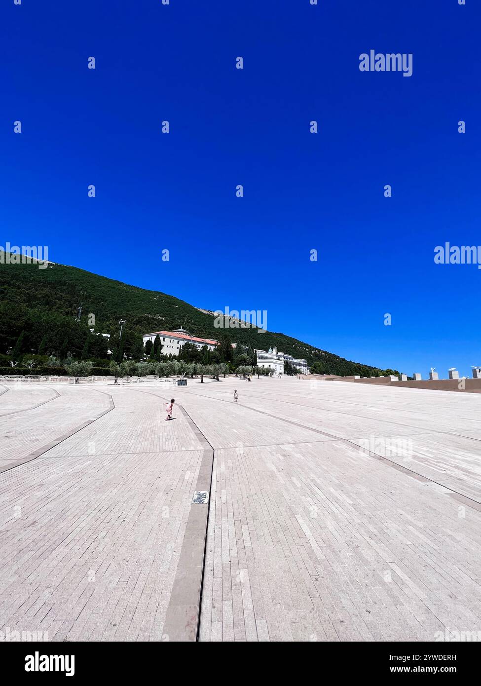 An expansive empty plaza stretches under a vibrant blue sky, with green ...