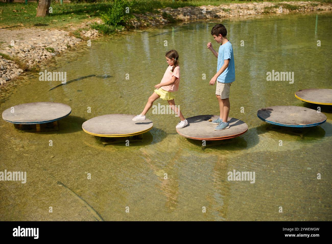 Two children balance on round stepping stones over a clear, shallow ...