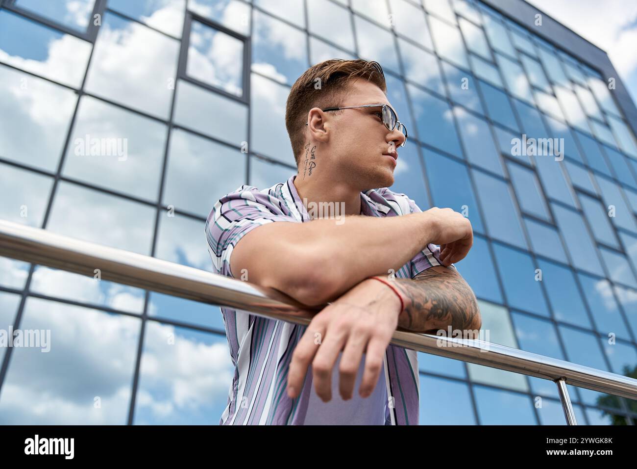 A young man with tattoos stands confidently by a sleek glass building ...