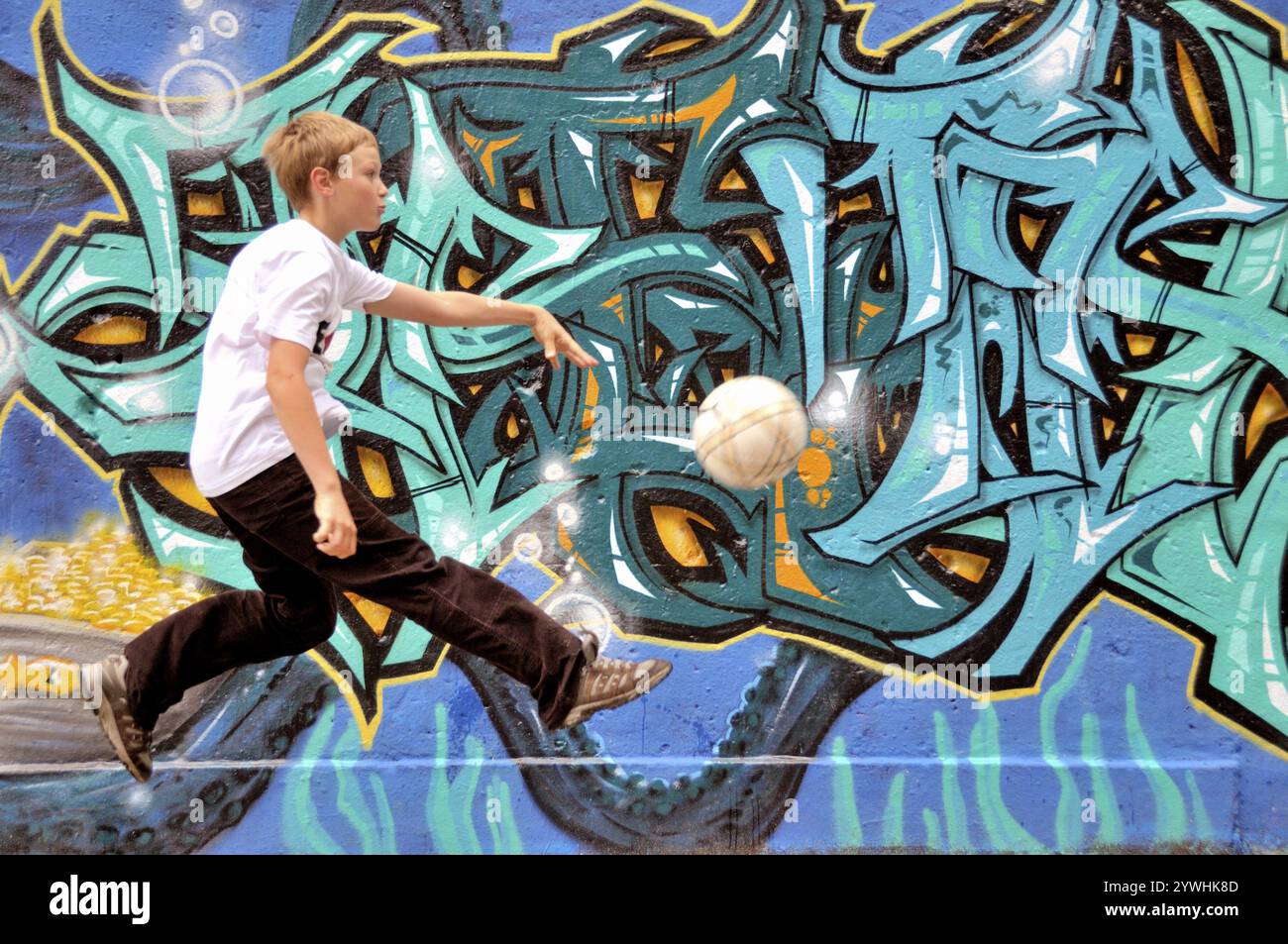 A ten-year-old boy plays with his football in front of a graffiti wall ...