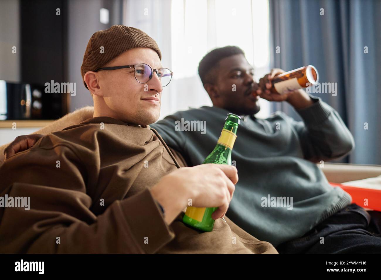 Portrait of two young men drinking beer while sitting on couch at home ...