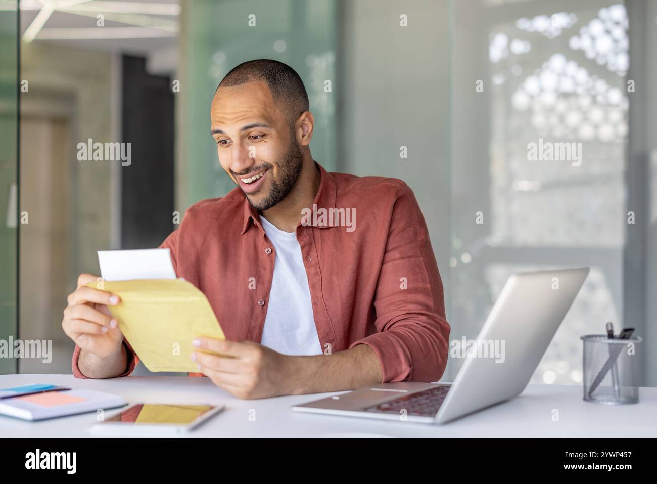 A cheerful man opening and reading an envelope at his office desk. He ...