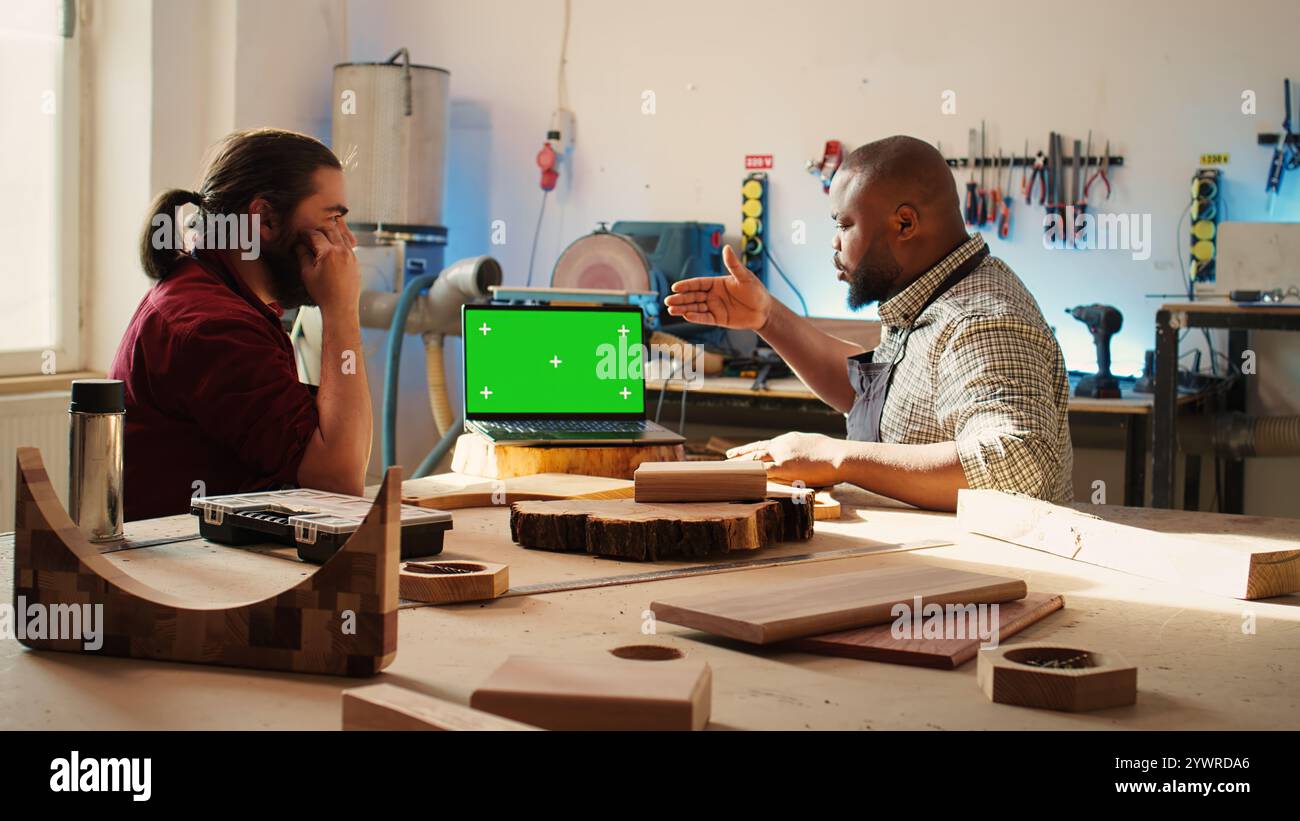 Woodworker looking over blueprints on green screen laptop in CAD software with apprentice ...