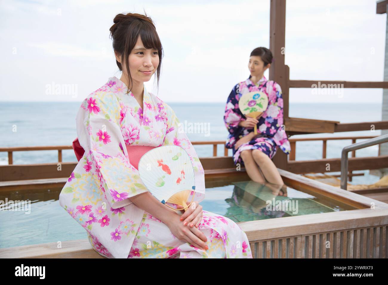 Two young women in yukata sitting on the edge of the open-air bath ...