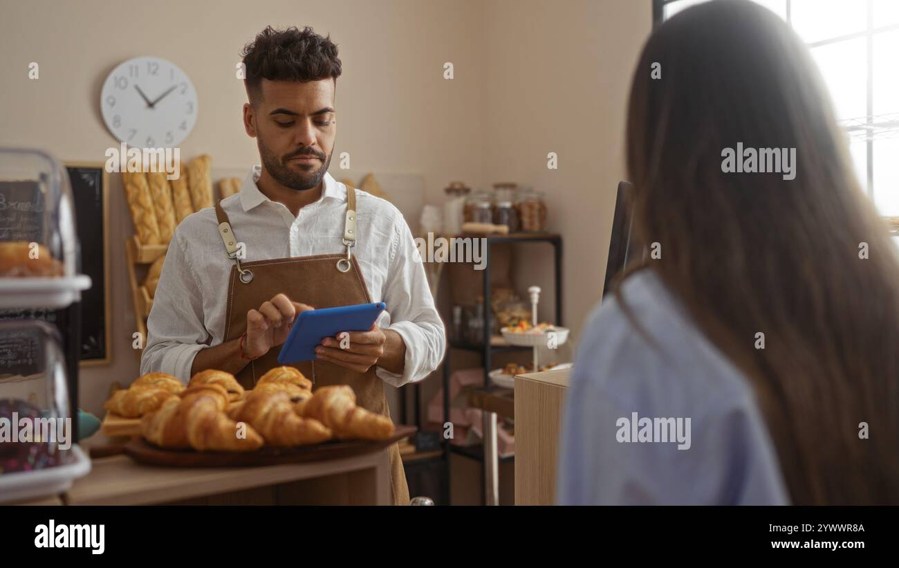Man in apron using tablet across countertop with pastries while woman waits in bakery interior ...