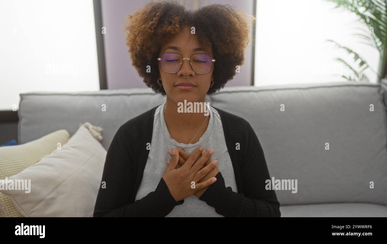 Woman meditating with eyes closed and hands on chest sitting on a sofa ...