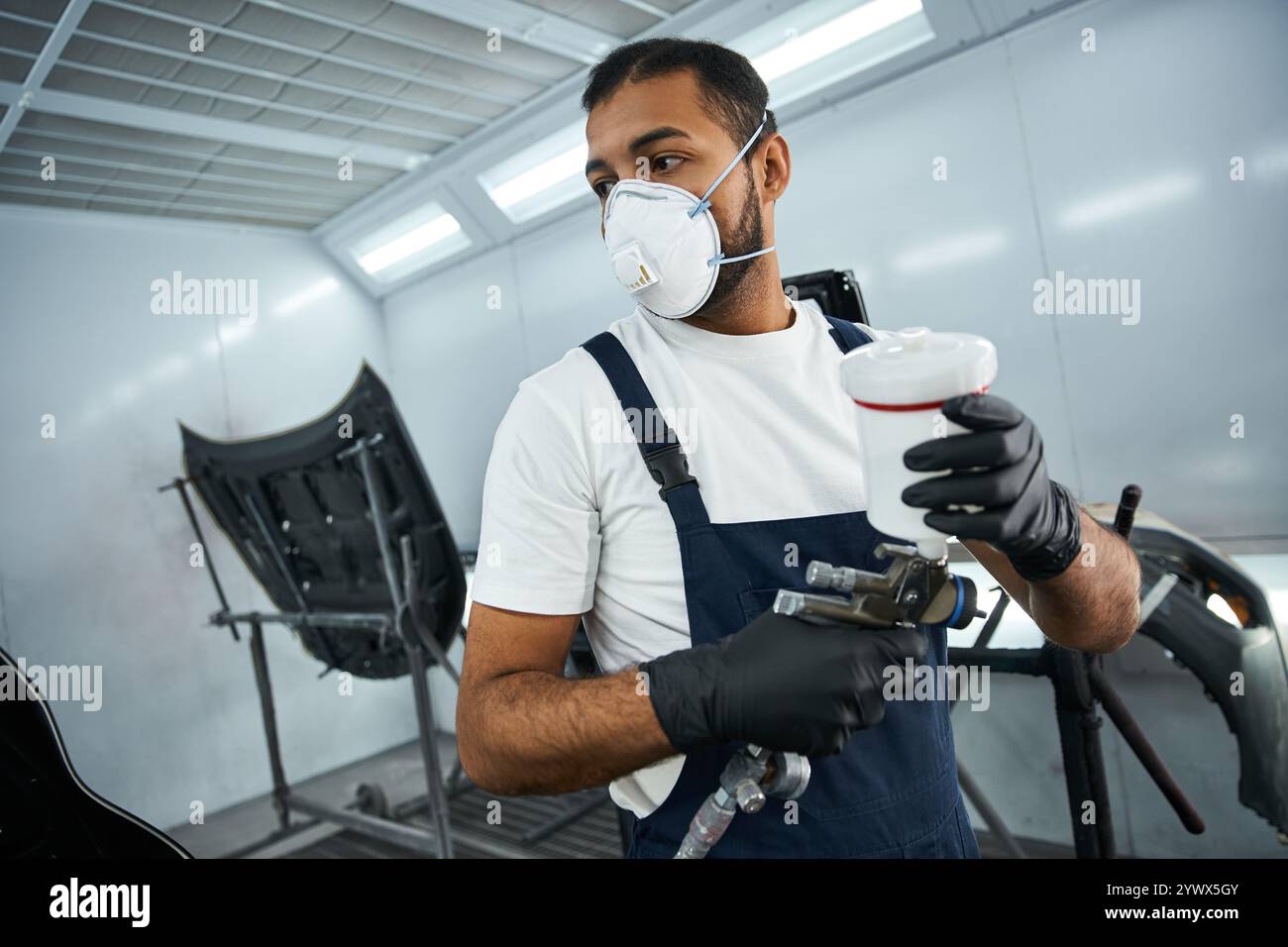 A young mechanic in work gear readies a spray gun for painting in the ...
