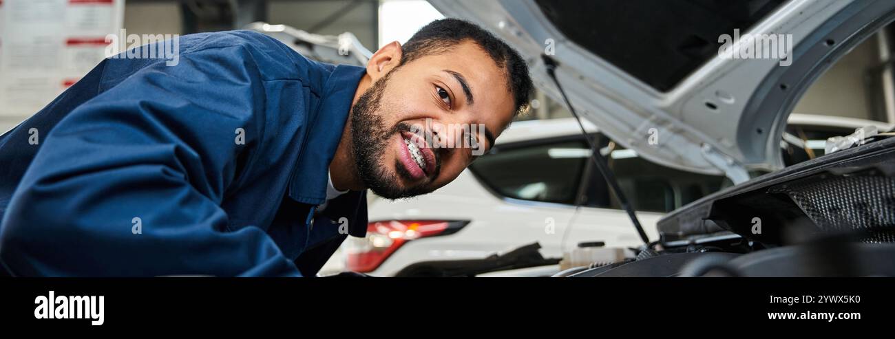 A young mechanic smiles while inspecting an engine under the hood in a ...