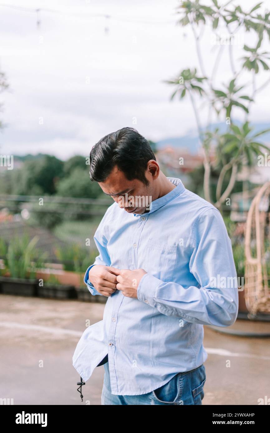 A Southeast Asian man adjusting his shirt button, focusing on the task ...