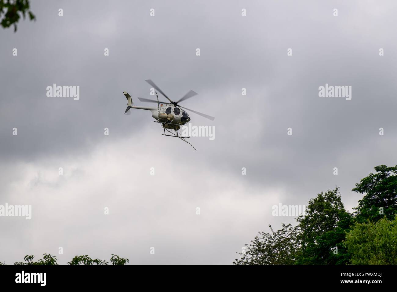 A small helicopter flying against a cloudy sky, with trees visible ...
