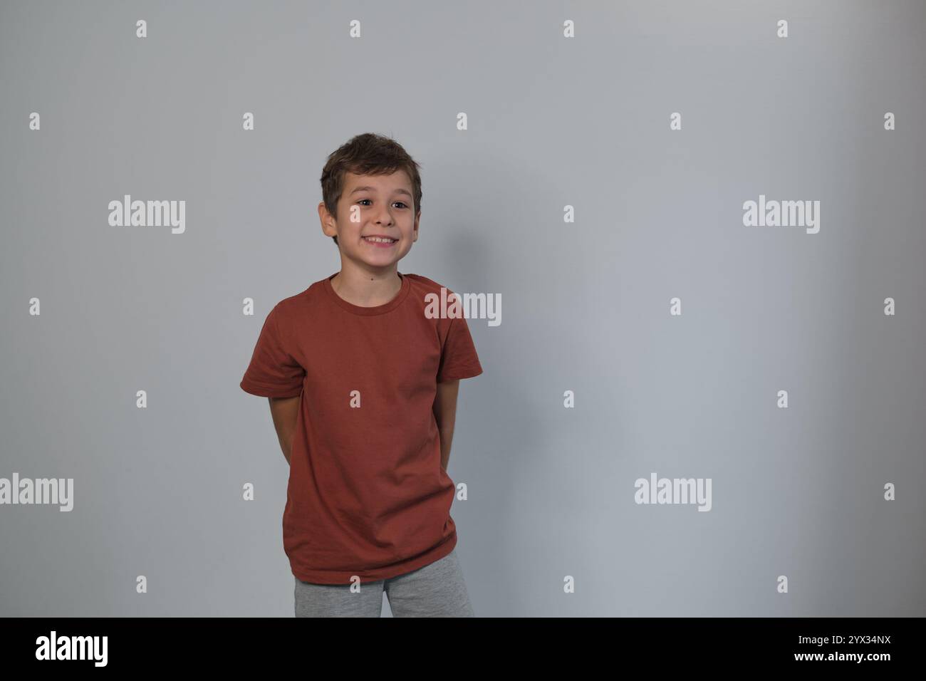 A boy in a rust tee and sweatpants stands against a light grey ...