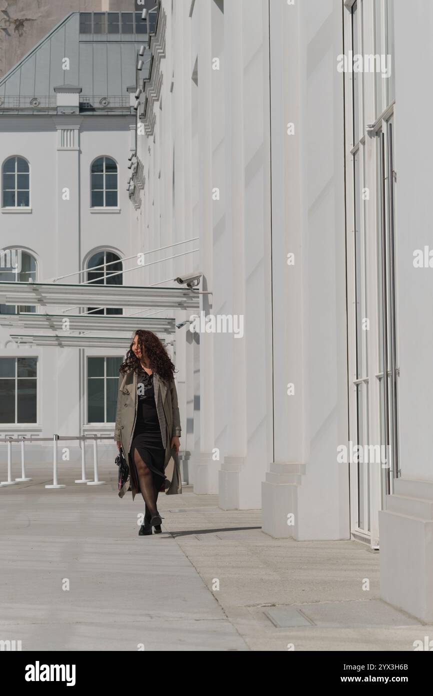 Woman dressed in a trench coat, walking by a sleek white building ...
