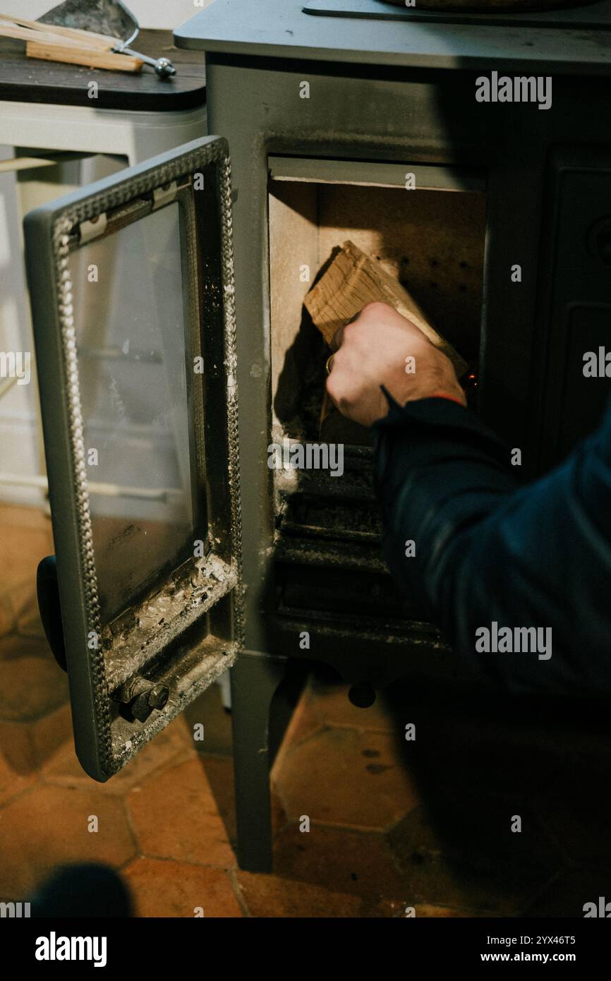 A man is putting wood into a stove. The stove is open and the wood is ...