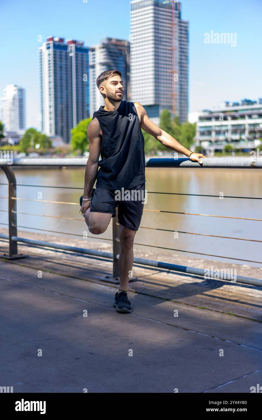 Fit young man engaging in flexibility exercises along a city canal ...