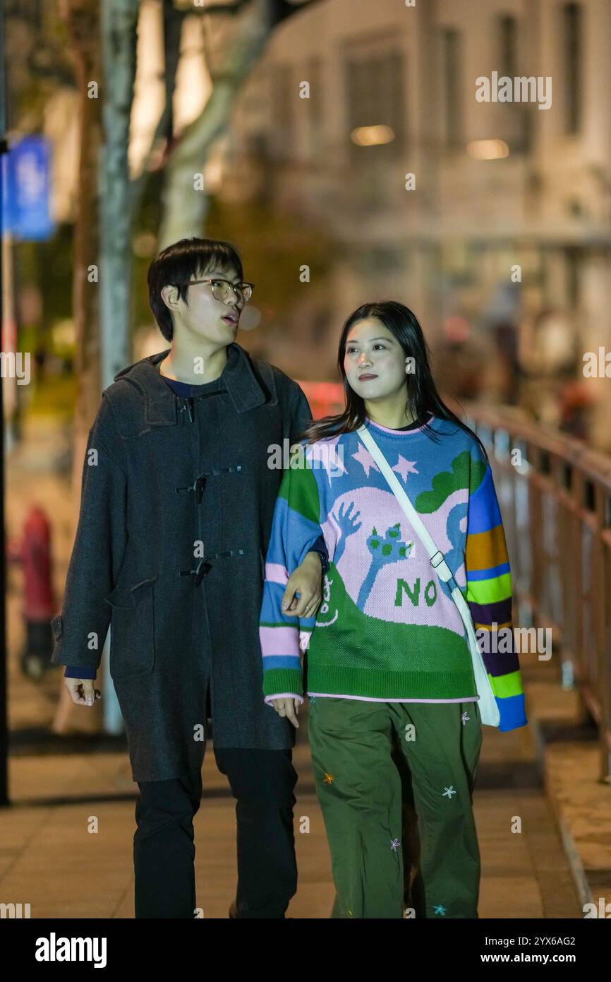A young Chinese couple in their 20s walking along a riverside sidewalk ...