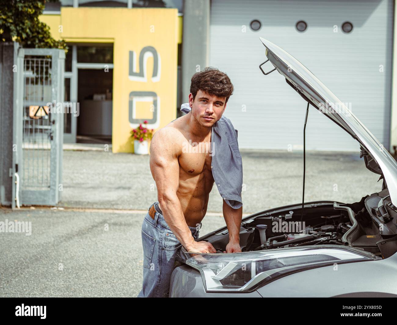 A young man with a muscular build examines the engine of a car with the ...