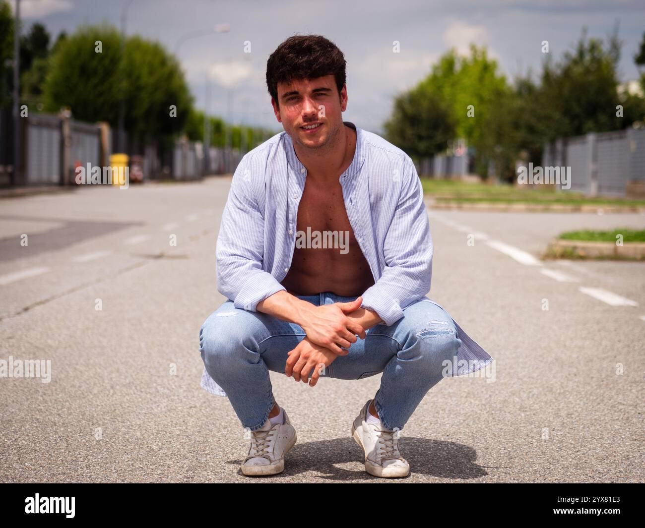 A young man squats on a deserted street, wearing a light shirt and ...