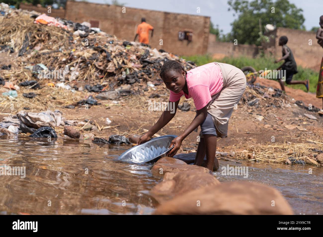Young African village girl washes dishes on the edge of a dirty river ...