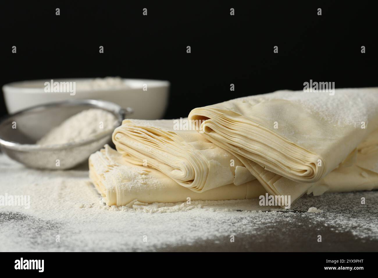 Raw puff pastry dough and flour on dark table against black background ...