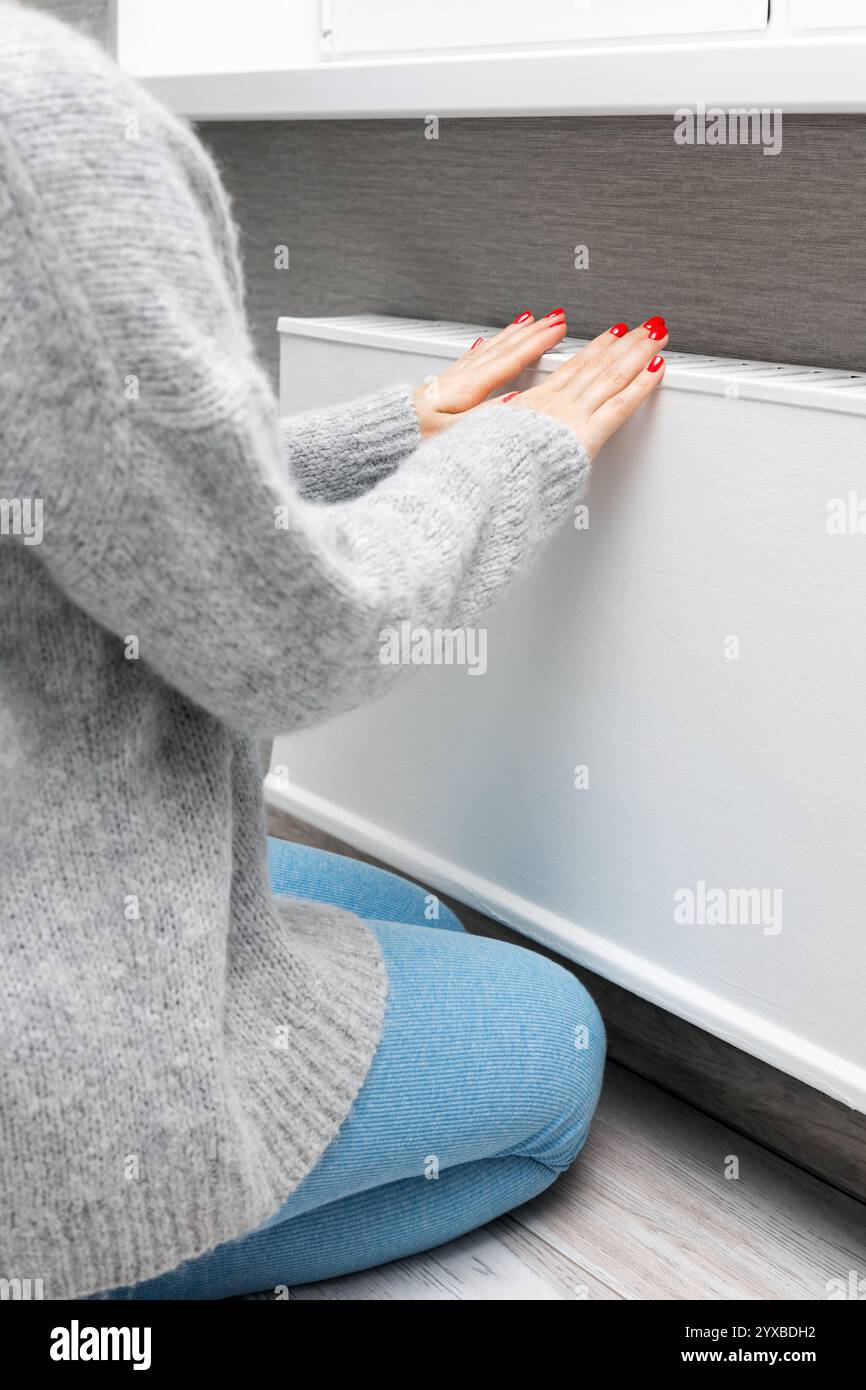 A girl sits near a heating radiator and warms her hands, vertical photo ...