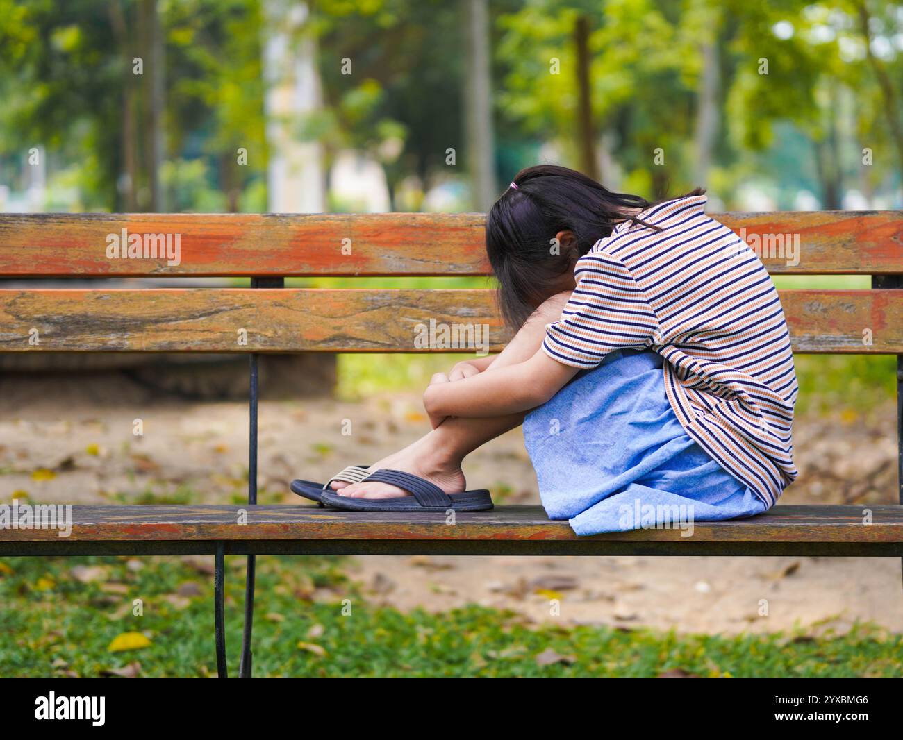 A sad little girl is sitting alone on bench in park. The little girl is ...