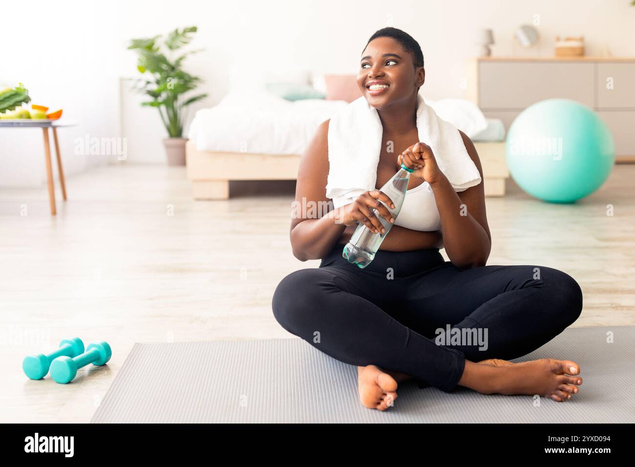 Plump black lady in sportswear sitting on sports mat with bottle of ...