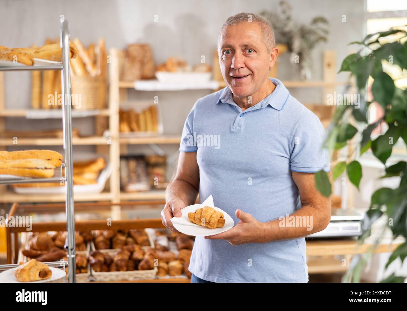 Senior male customer stands with croissant in hands near window of ...