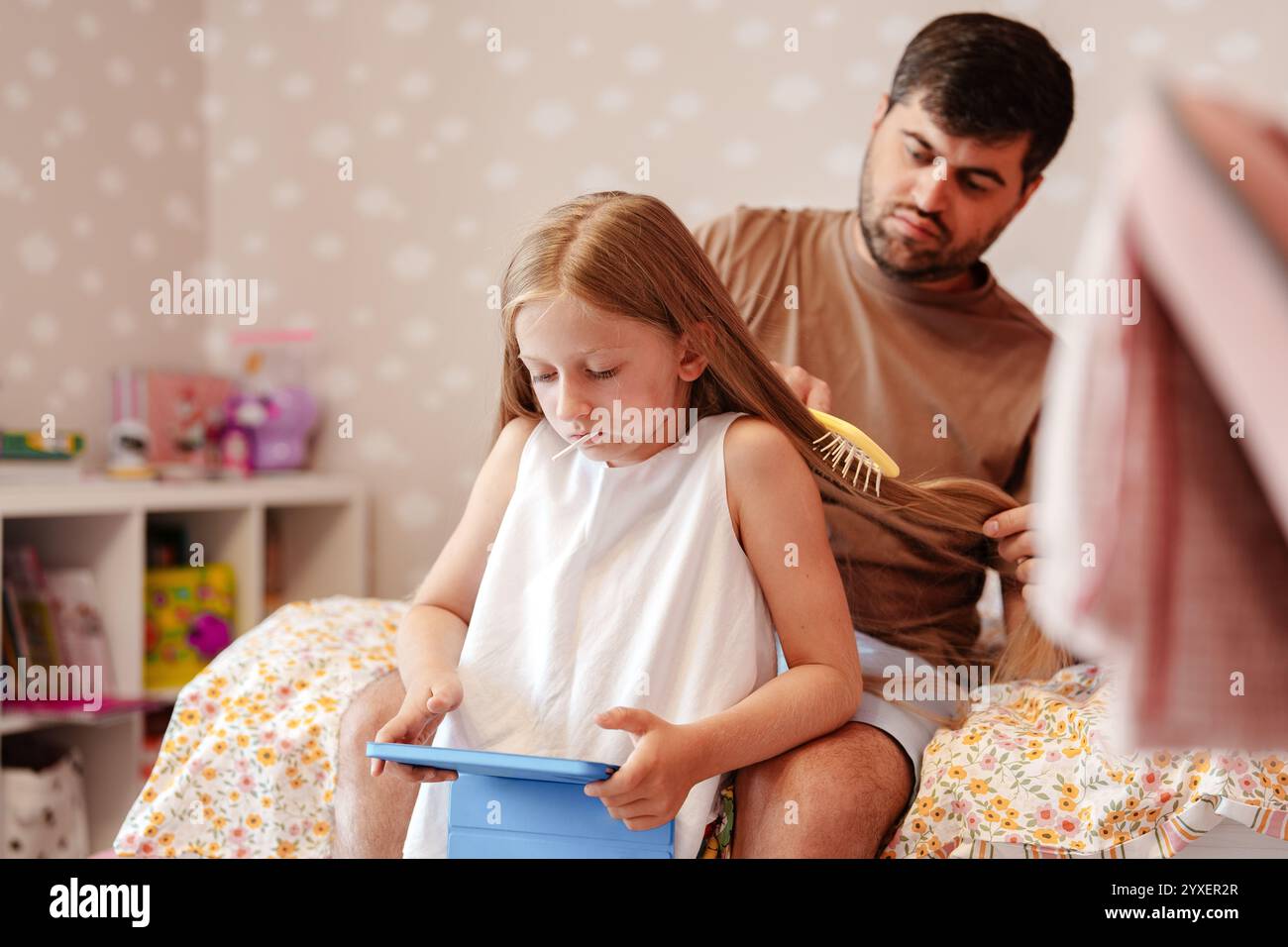 A father brushes his daughter's hair while she uses a tablet in her ...