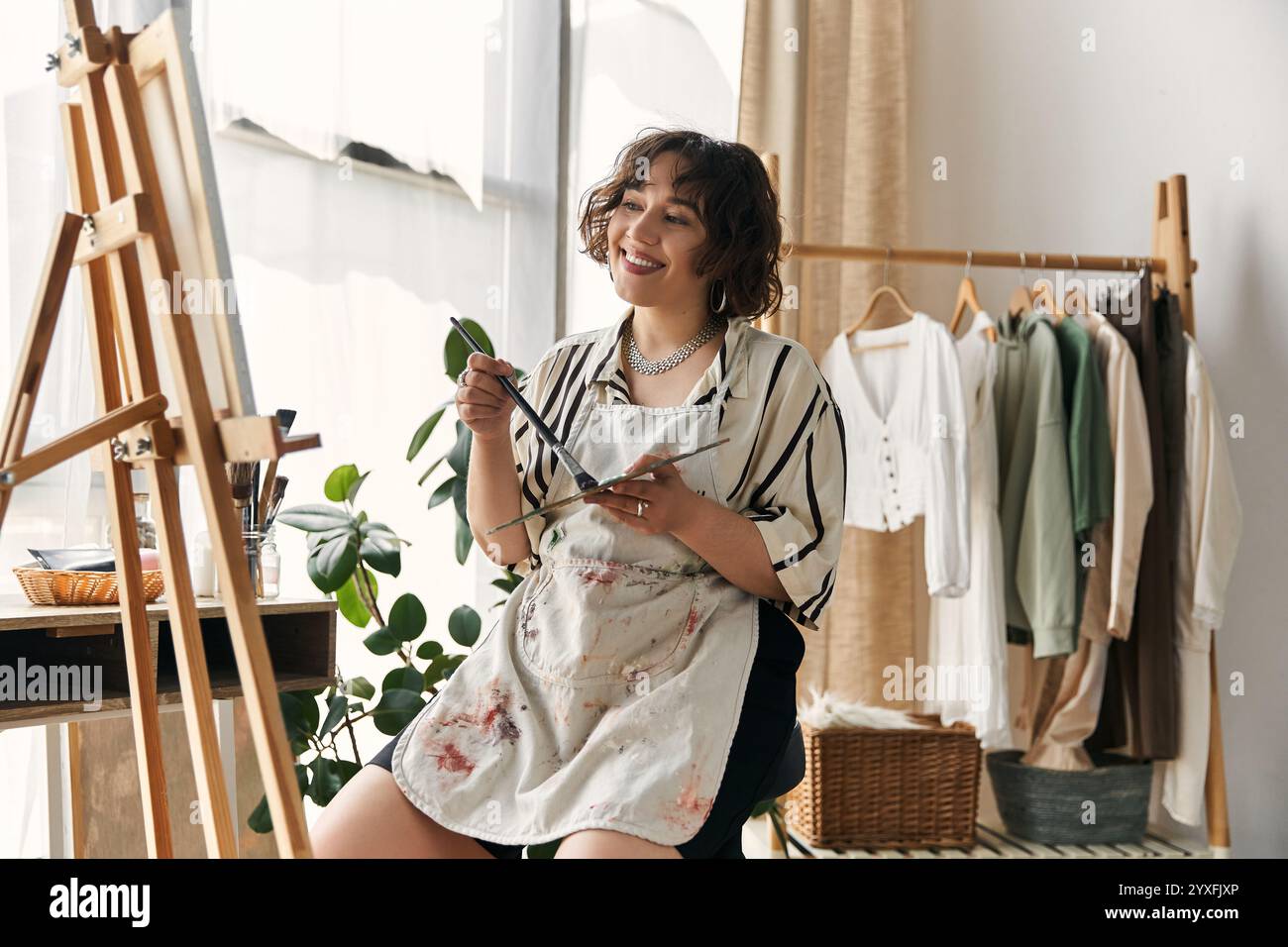 A joyful young woman with curly hair paints art on an easel in her ...