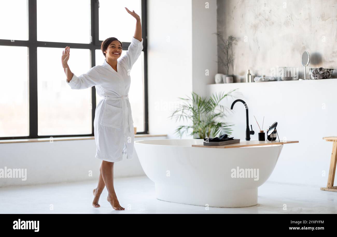 Woman in white robe joyfully dancing in modern bathroom by large window ...