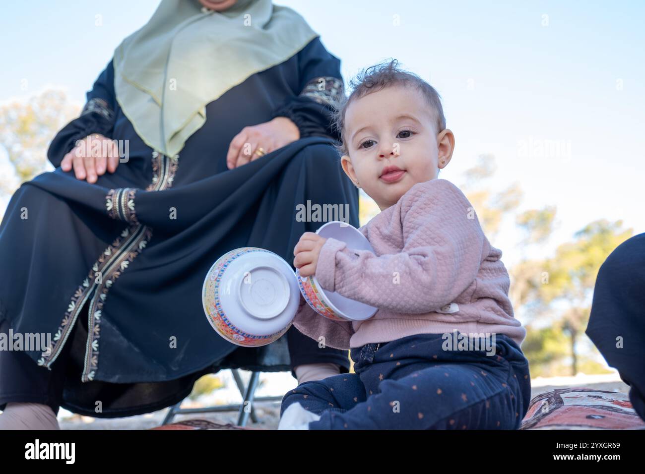 A young child sitting on a mat outdoors, holding bowls, with a family ...