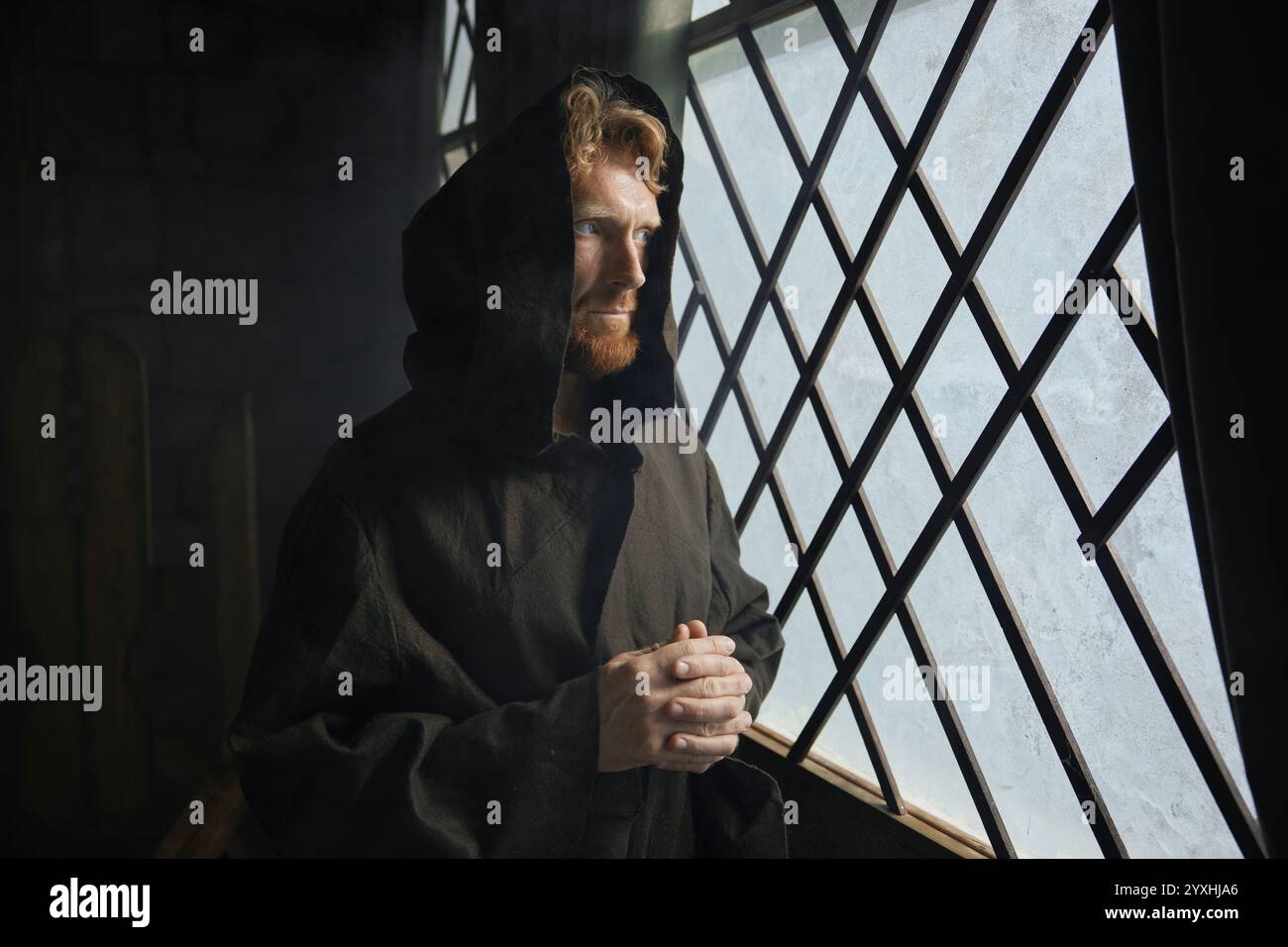 Pensive monk in medieval costume with hood looking through window of ...