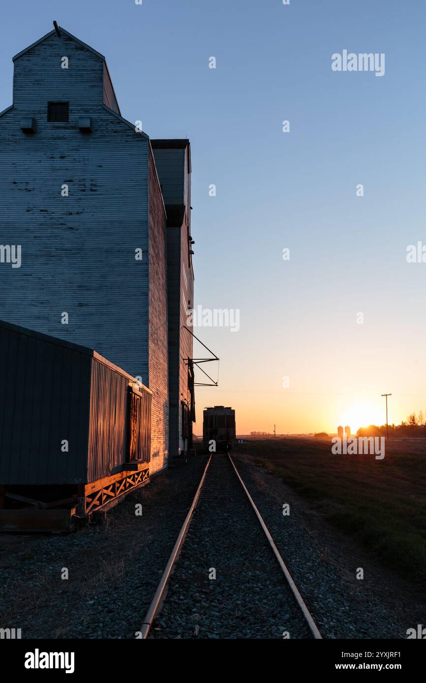 A train is traveling down a track next to a grain silo. The sun is ...