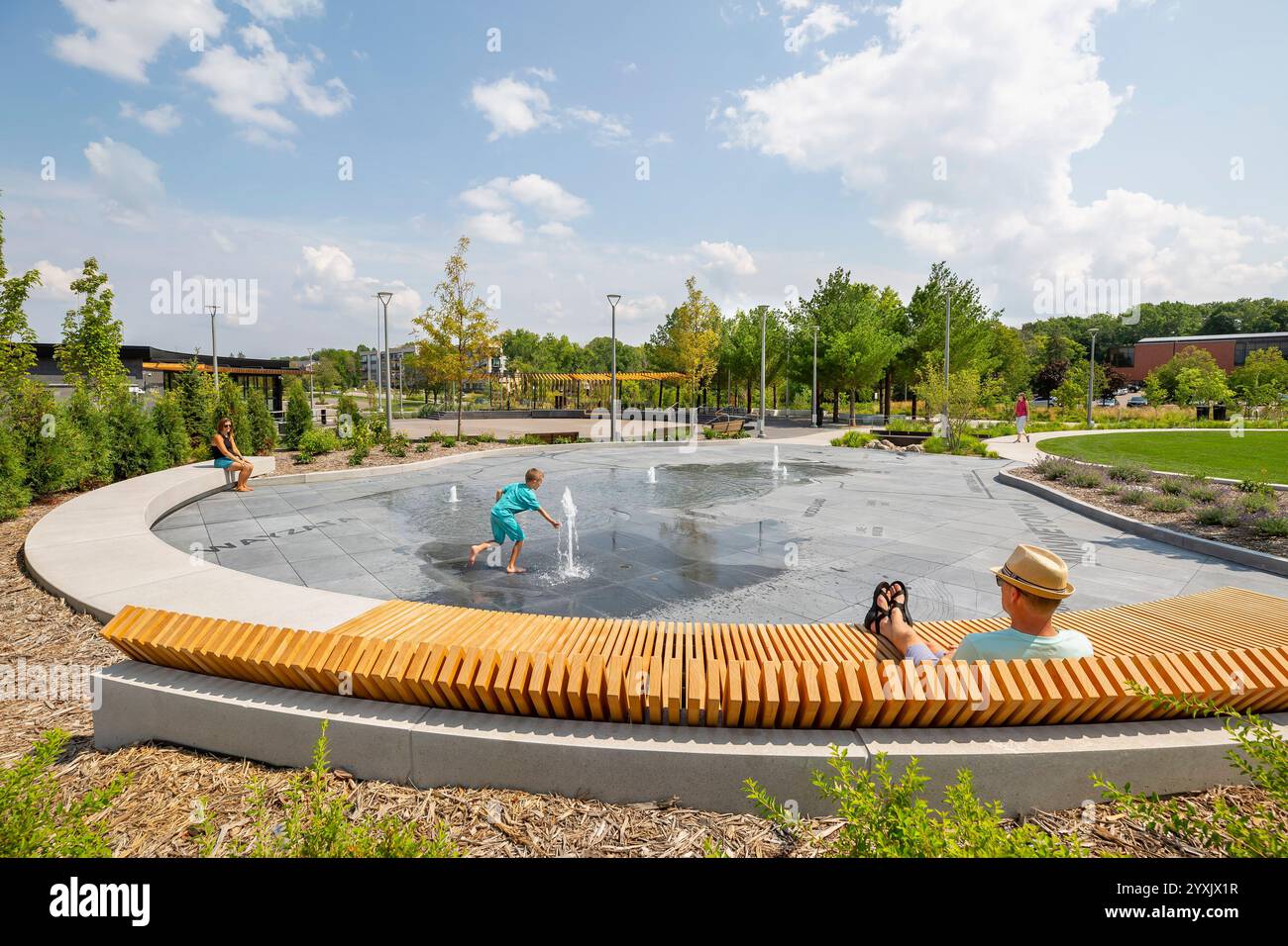 A child plays with on a splash pad for an interactive water feature as two adults look on in ...